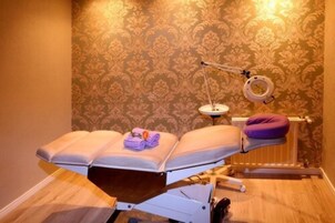 Sala de tratamentos com mesa de massagem, aparelho de massagem para os pés e uma pequena mesa com toalhas.