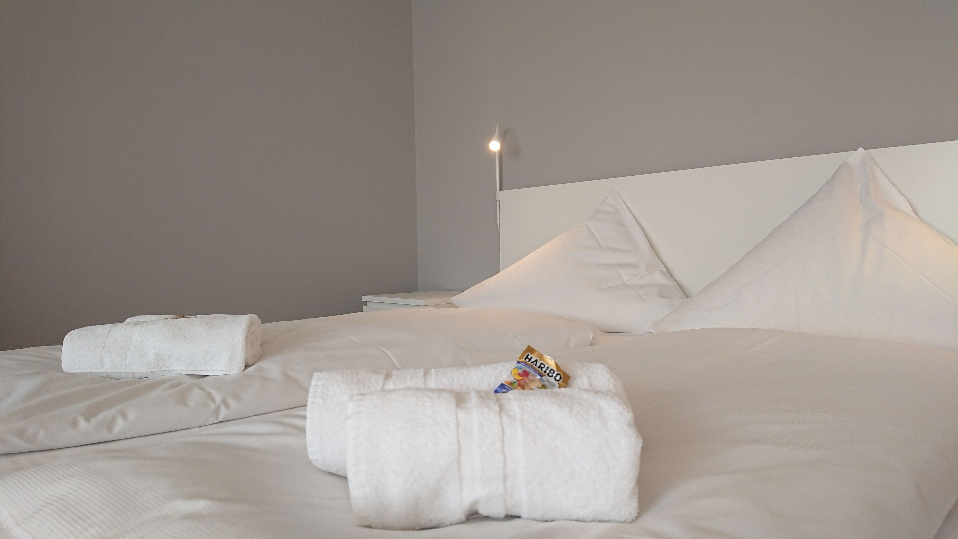 Quarto de hotel com duas camas, roupa de cama branca e uma barra de caramelos Haribo em uma das camas.