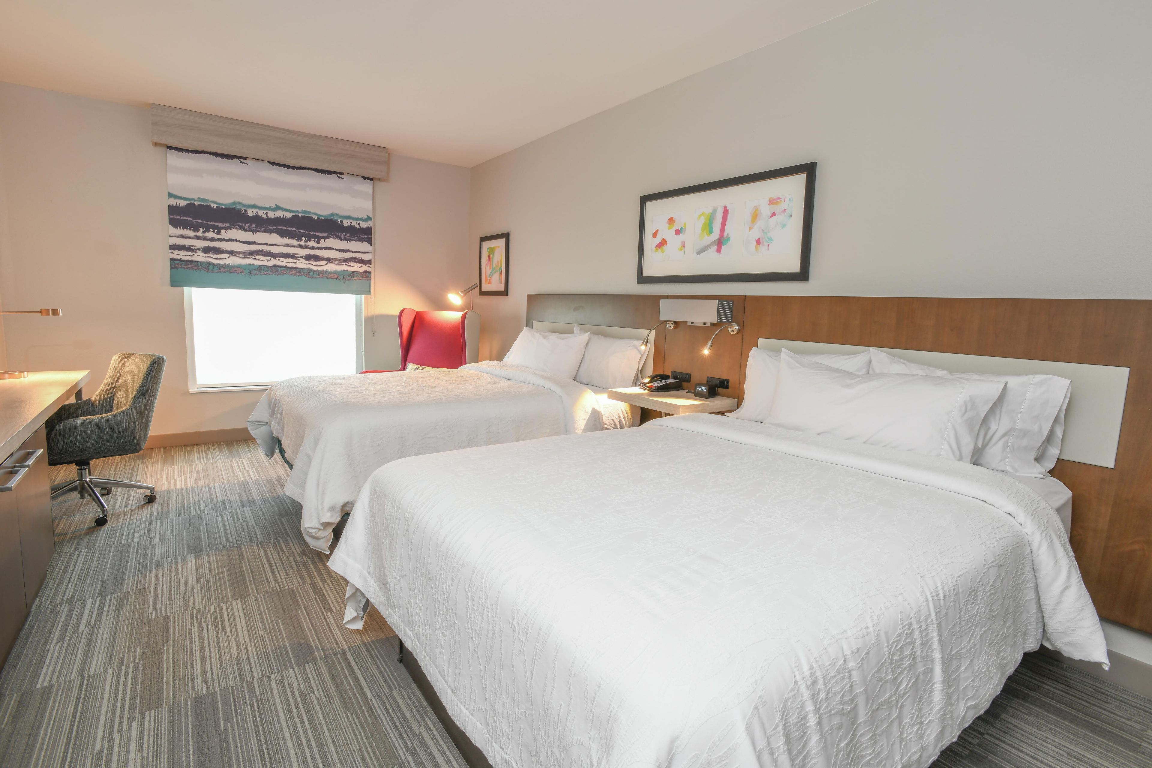 Quarto de hotel com duas camas, uma escrivaninha, uma cadeira e uma janela com vista.