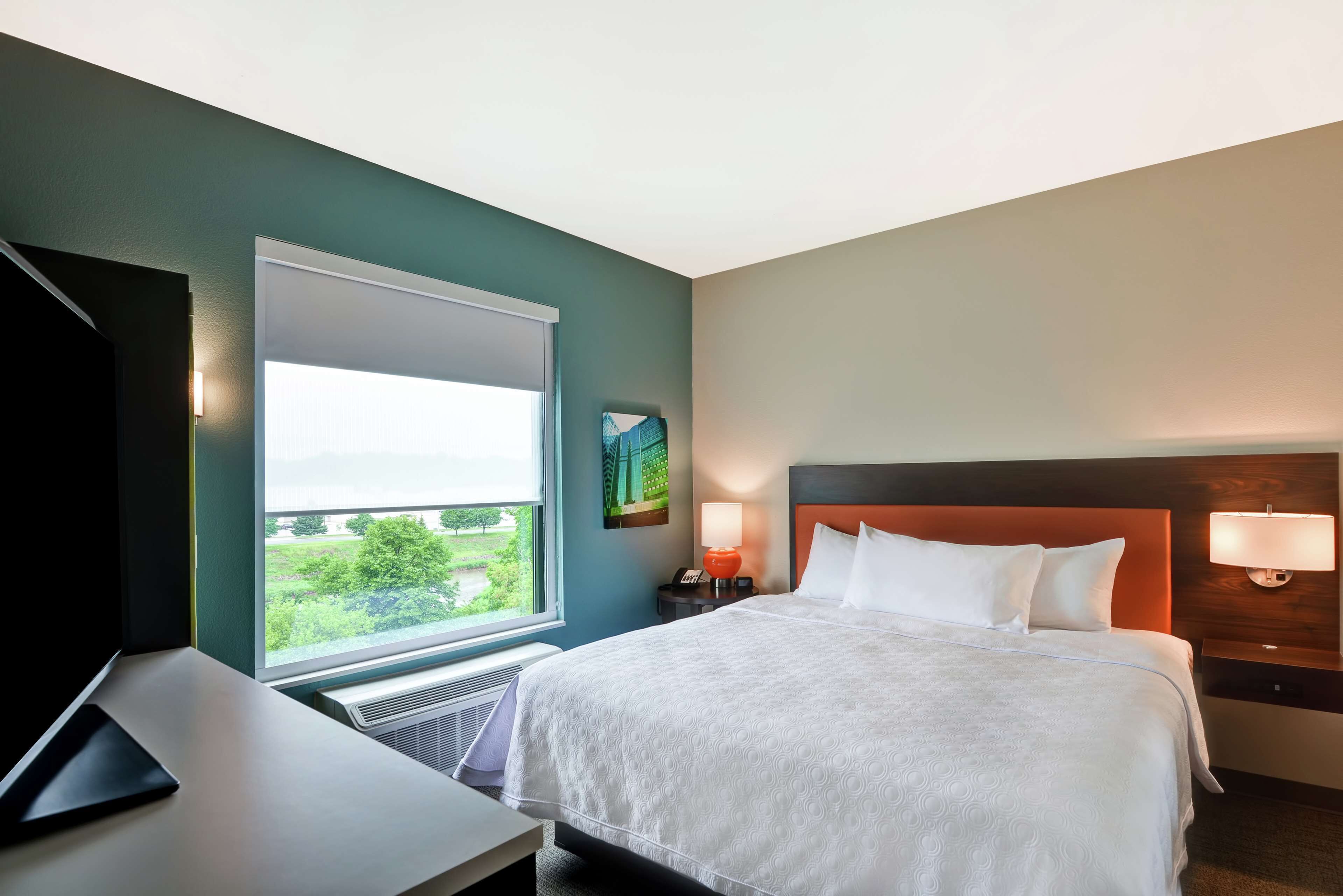 Quarto de hotel com uma cama grande, uma escrivaninha com um laptop, uma janela com vista para áreas verdes e uma mesa de cabeceira com abajur.