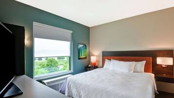 Quarto de hotel com uma cama grande, uma escrivaninha com um laptop, uma janela com vista para áreas verdes e uma mesa de cabeceira com abajur.