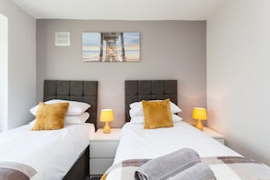 Quarto de hotel com duas camas, um quadro de um cais, uma parede branca e uma janela.