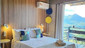 Quarto com teto de madeira, cama com roupa de cama branca, uma mesinha e vista para montanhas.