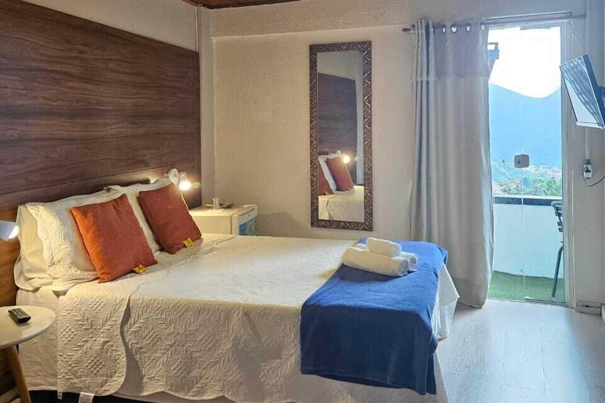 Quarto com teto de madeira, cama com roupa de cama branca e azul, mesa de cabeceira, espelho e vista para o exterior.