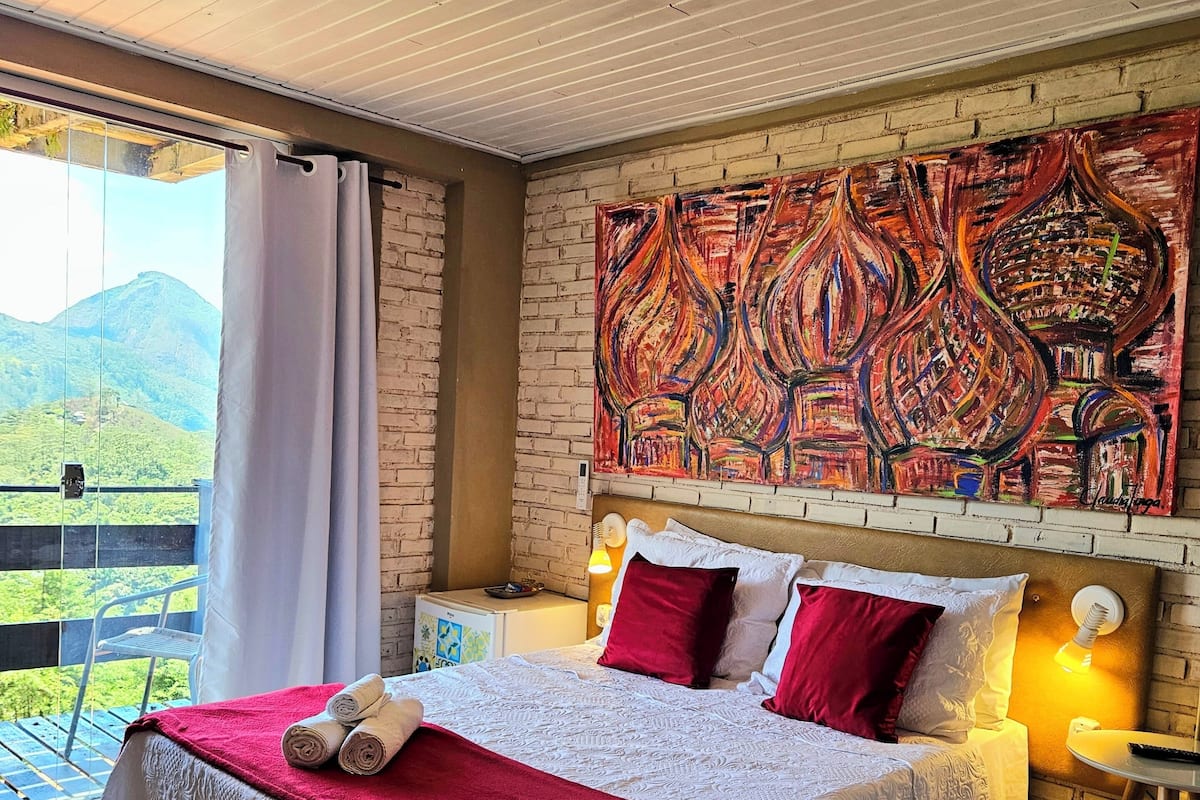 Quarto com cama, janela grande, parede de tijolos e uma obra de arte colorida.
