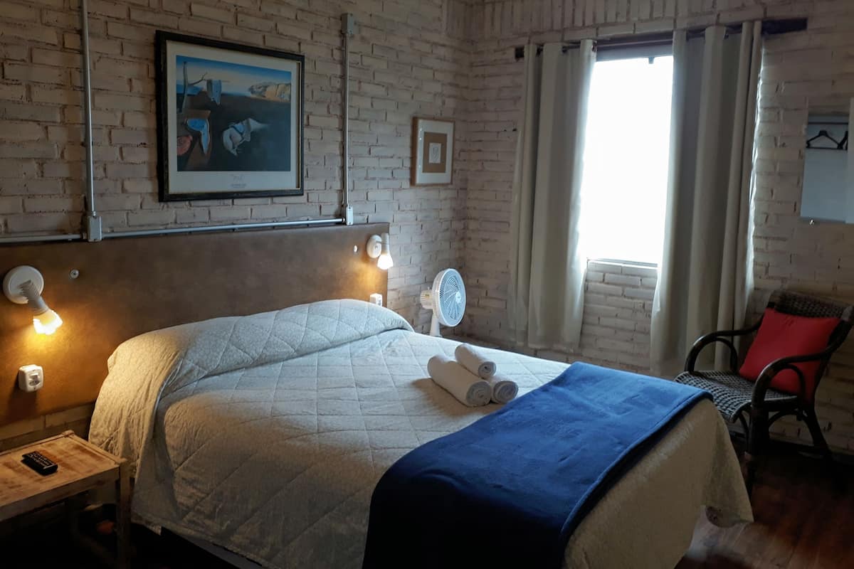 Quarto de hotel com parede de tijolos, cama com colcha, mesa de cabeceira com controle remoto, ventilador e cadeira com almofada vermelha.