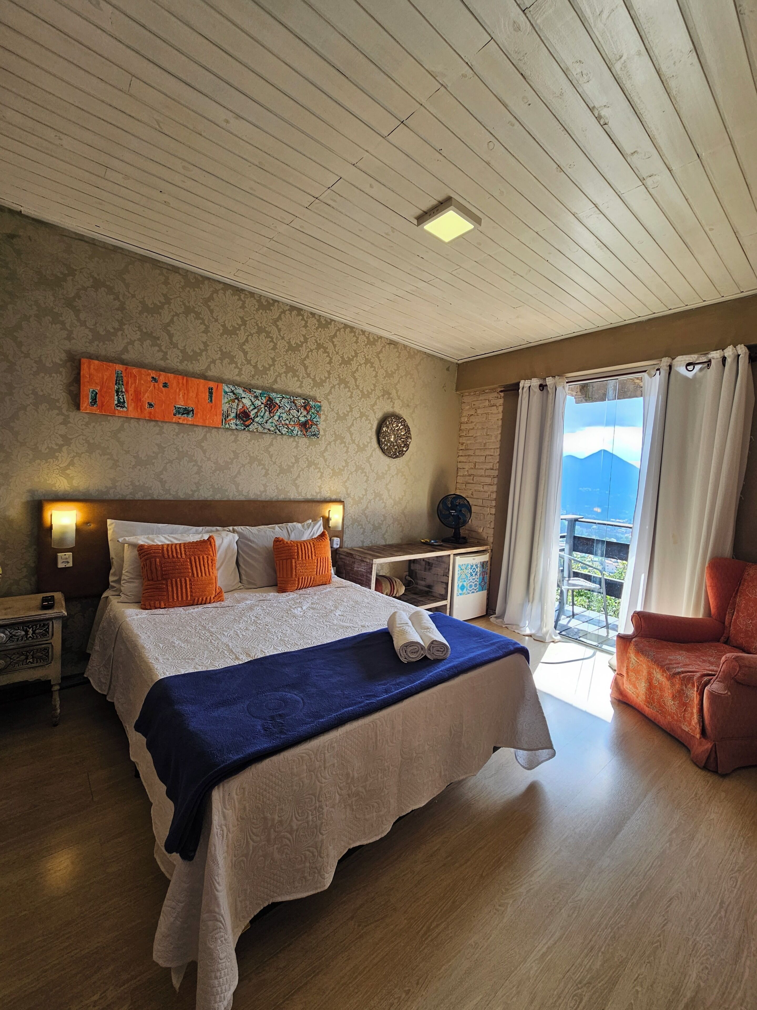 Quarto com uma cama grande, mesa de cabeceira, cadeira, janela com cortinas e vista para o exterior.
