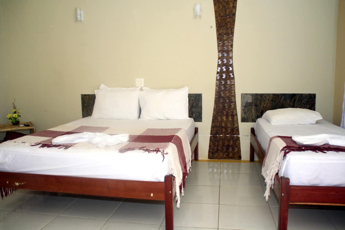 Quarto de hotel com duas camas, cada uma com roupa de cama branca e cabeceira marrom. Há uma pequena mesa com um vaso de flores e um candeeiro fixado na parede.
