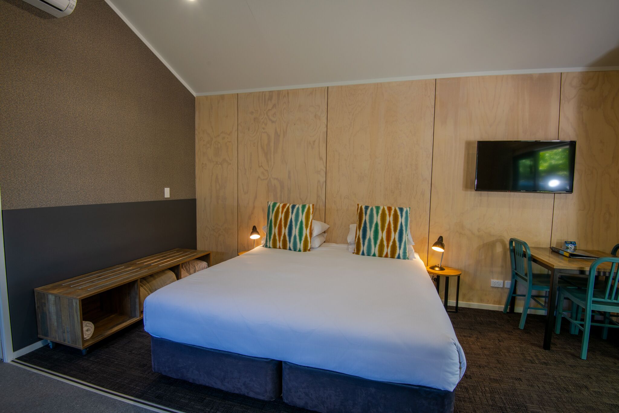 Quarto de hotel com uma cama grande, paredes de painel de madeira, televisão, secretária com cadeiras e um banco.