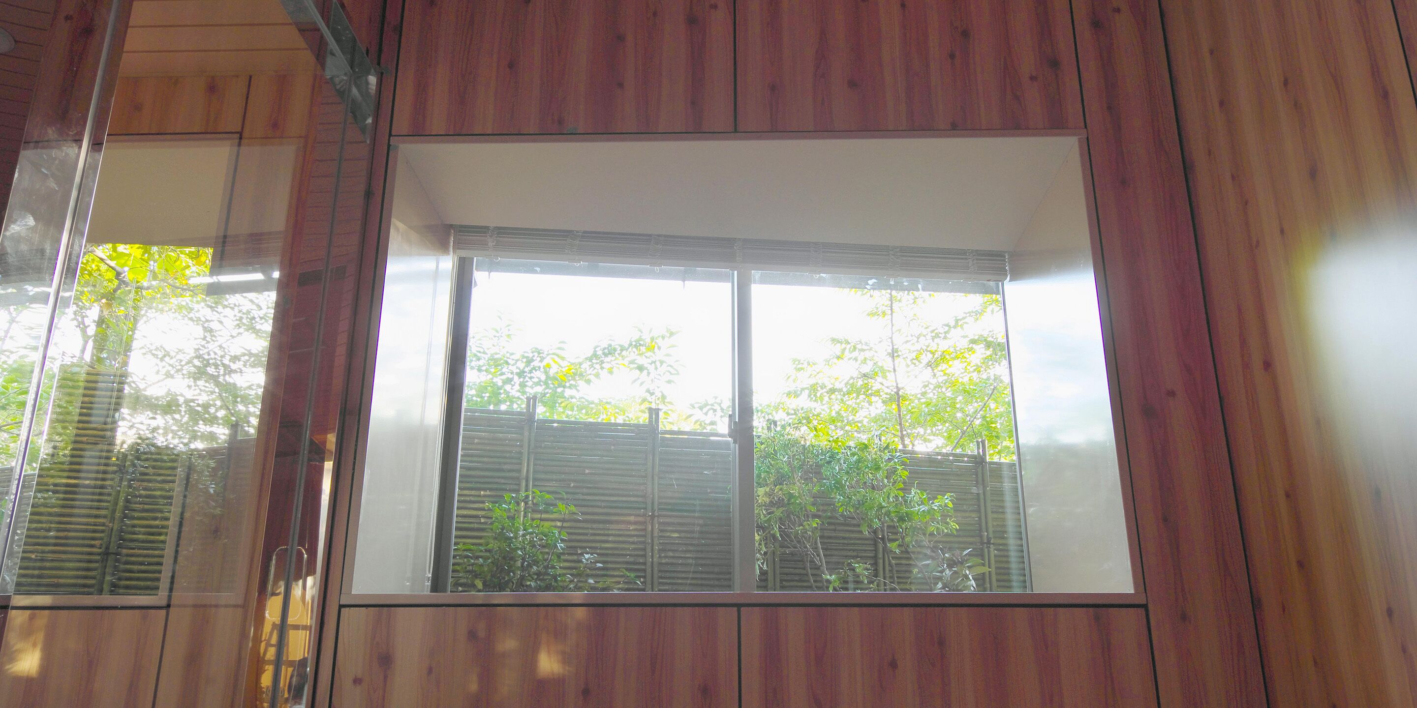 Quarto com painéis de madeira, uma janela grande com persianas e vista para uma cerca de bambu e árvores.