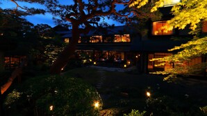 Um edifício tradicional japonês, com vários níveis, janelas iluminadas e um jardim com um caminho.