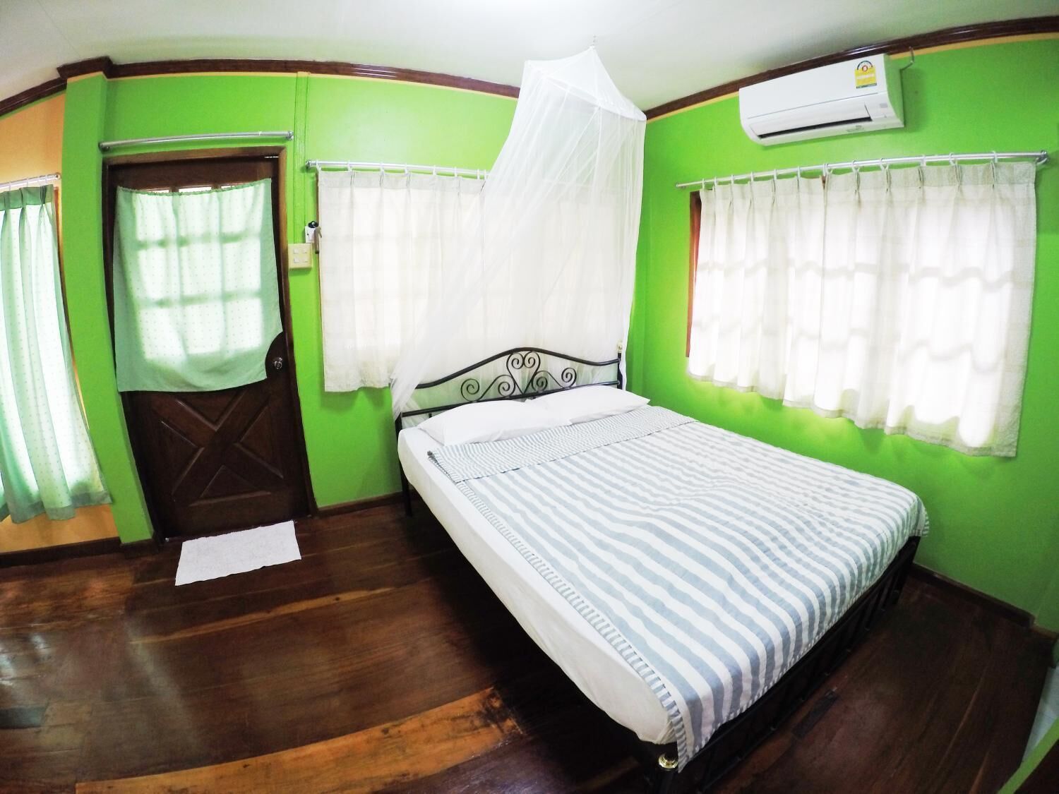Um quarto com cama, paredes verdes, assoalho de madeira, janela com cortinas brancas e um aparelho de ar condicionado.