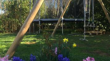 Um parquinho com uma ponte de corda e balanços, cercado por vegetação e flores.