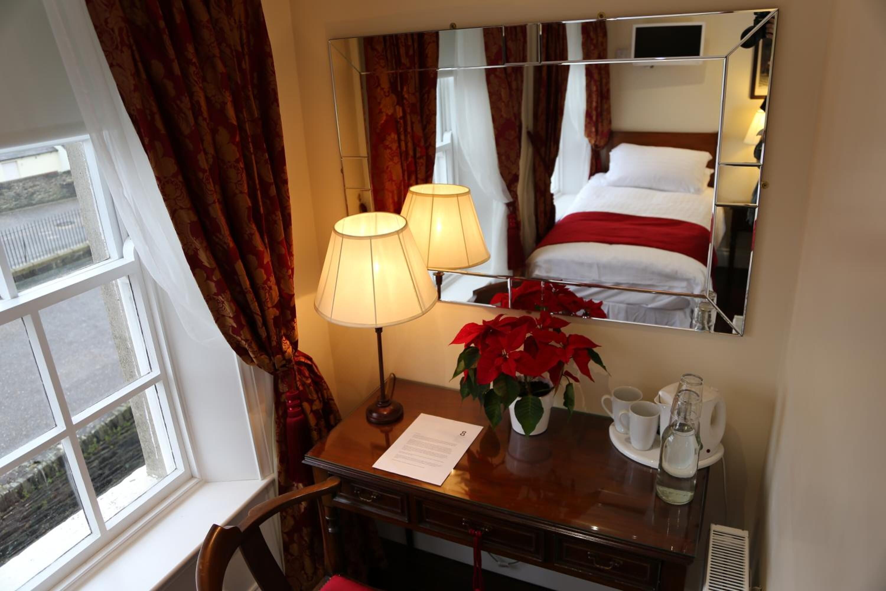 Quarto de hotel com cama, duas luminárias de cabeceira, escrivaninha com espelho, um vaso com flores vermelhas e janela com cortinas.
