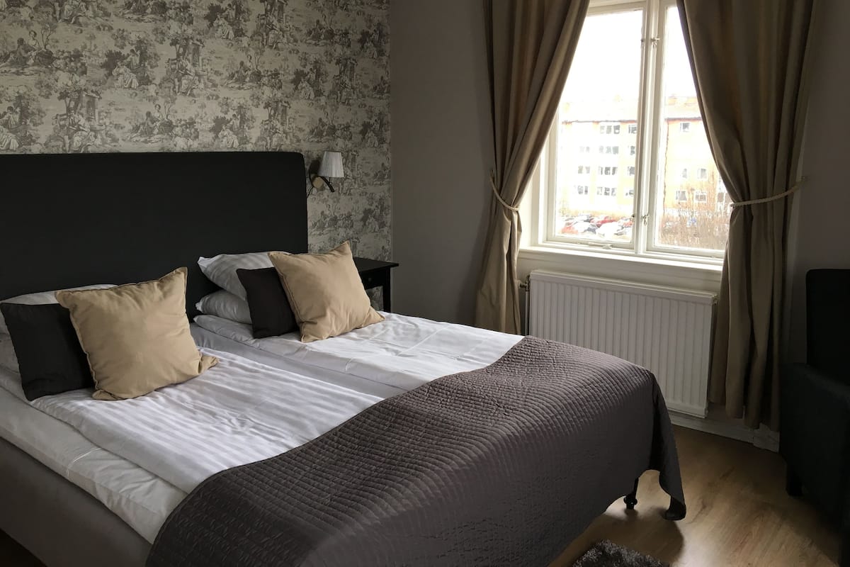 Cama bem arrumada com cabeceira escura, mesa de cabeceira com abajur e janela com cortinas.