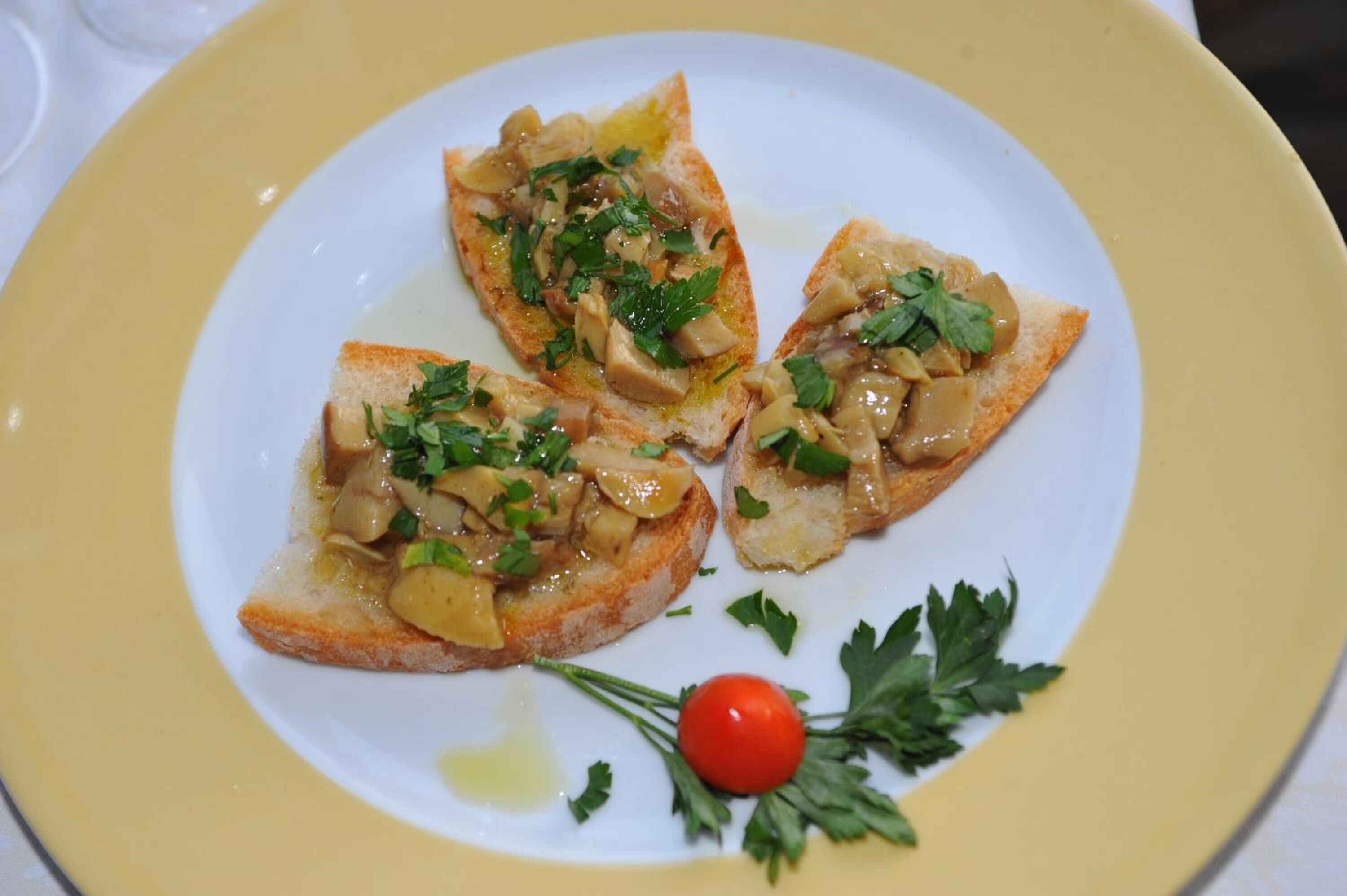 Um prato com três bruschettas cobertas de cogumelos e salsa, decorado com um tomate cereja e um ramo de salsa.