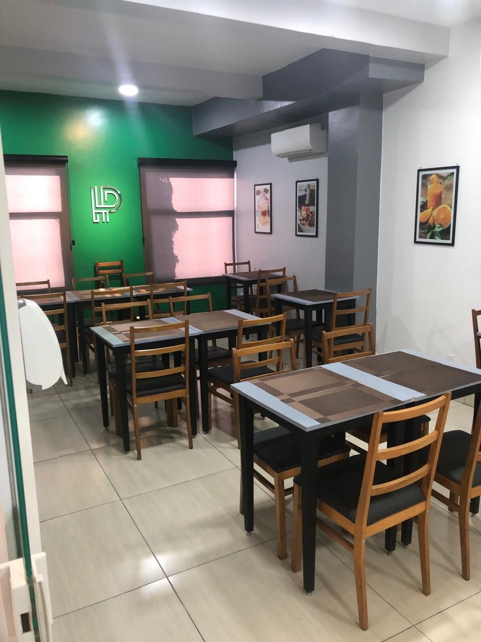 Uma sala de jantar com várias mesas e cadeiras, um muro verde de destaque e quadros nas paredes.
