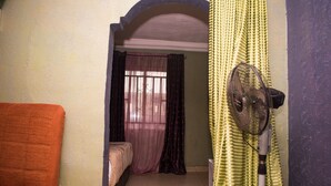 Um corredor estreito com uma cama, uma cadeira e um ventilador.