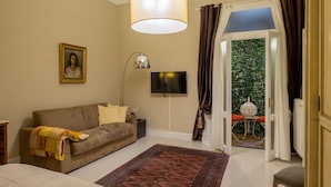 Uma sala de estar com sofá, televisão, abajur, tapete estampado e porta que dá para uma varanda com vegetação.