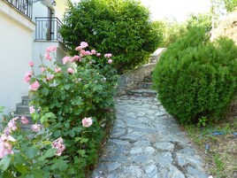 Uma trilha de pedra ladeada por vegetação exuberante e rosas cor-de-rosa, que leva a uma escadaria de pedra.