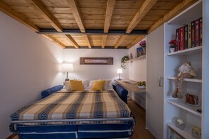 Quarto com teto de madeira, cama com roupa de cama estampada, duas luminárias de cabeceira, mesa de cabeceira e estante com livros e objetos decorativos.