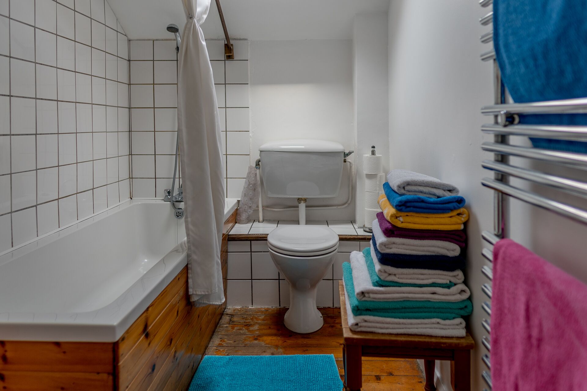 Uma casa de banho pequena com sanita branca, banheira e uma pilha de toalhas numa estante.