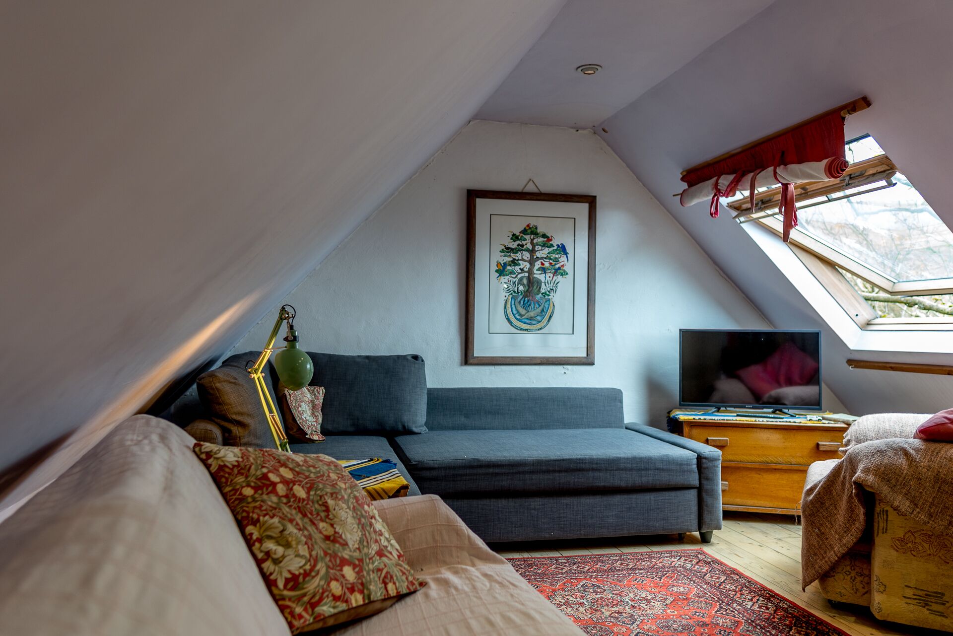 Quarto de sótão acolhedor com um sofá azul, uma obra de arte emoldurada, uma televisão e um claraboia.