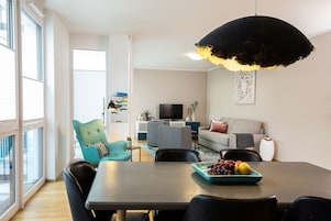 Uma sala de estar moderna com área de jantar, janelas amplas, luminária pendente preta e televisão de tela plana.