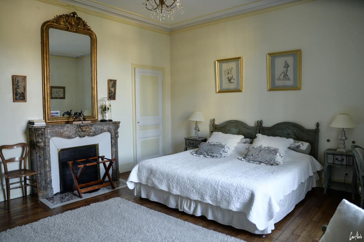 Um quarto com uma cama grande, lareira, espelho e quadros nas paredes.
