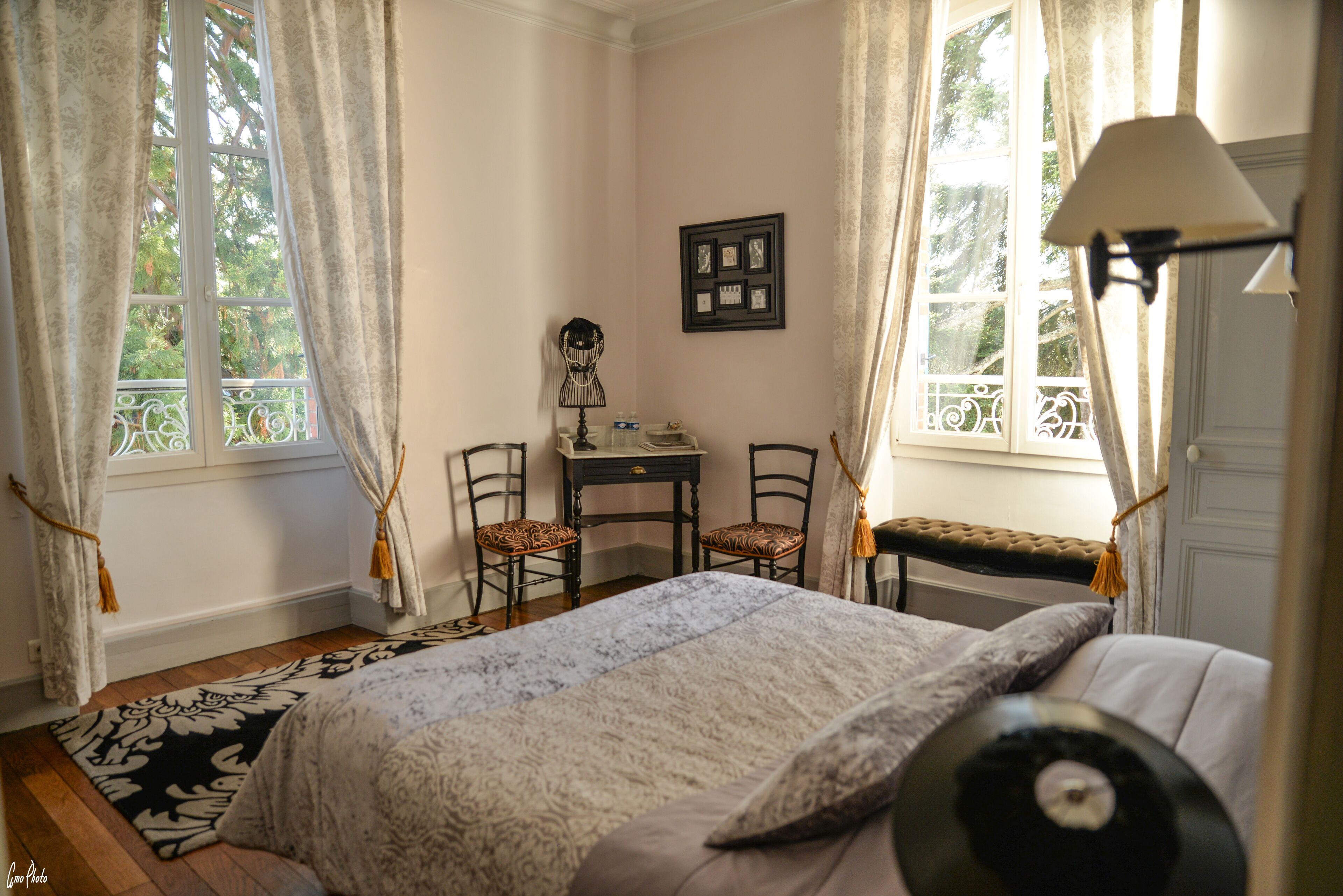 Quarto com cama, escrivaninha, duas cadeiras, abajur e janela com cortinas.
