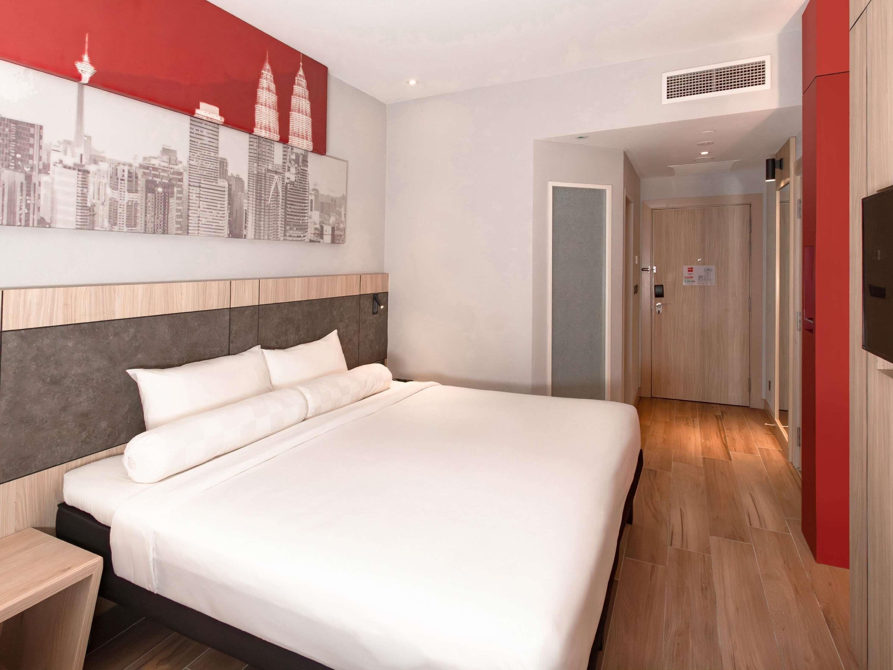 Quarto de hotel com uma cama grande, uma obra de arte com vista para a cidade na parede, piso de madeira e uma parede com detalhes em vermelho.