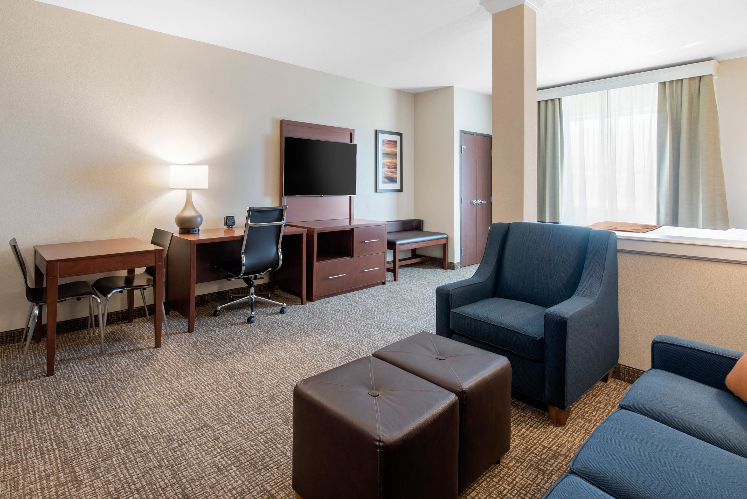 Quarto de hotel com escrivaninha, cadeira, televisão, banquinho e sofá.