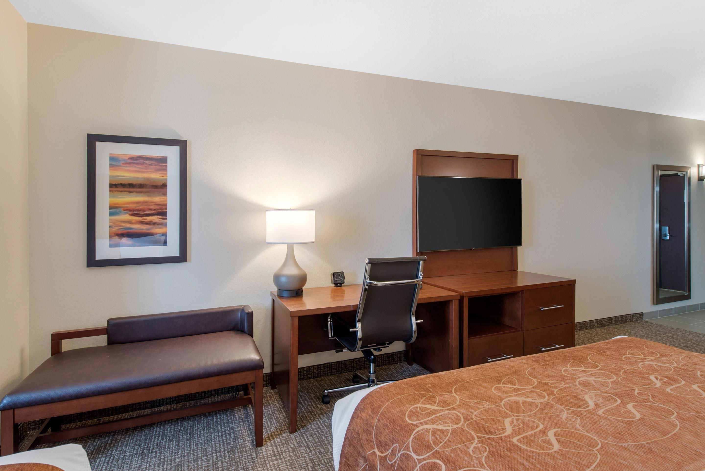 Quarto de hotel com uma cama, uma escrivaninha com cadeira, televisão, luminária e um quadro na parede.
