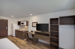 Quarto de hotel compacto com cozinha compacta, uma cama, escrivaninha com cadeira, televisão e prateleiras embutidas.