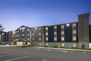 Um hotel moderno com uma placa chamativa que diz "Woodspring Suites". Possui um amplo estacionamento com vagas demarcadas e uma entrada bem iluminada.