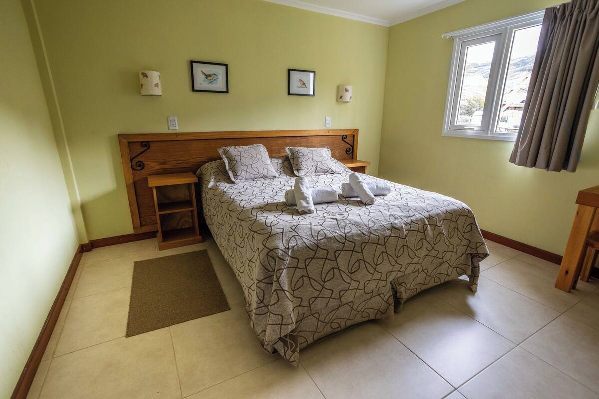 Quarto bem organizado com cama, mesas de cabeceira, janela com cortinas e arte pendurada na parede.
