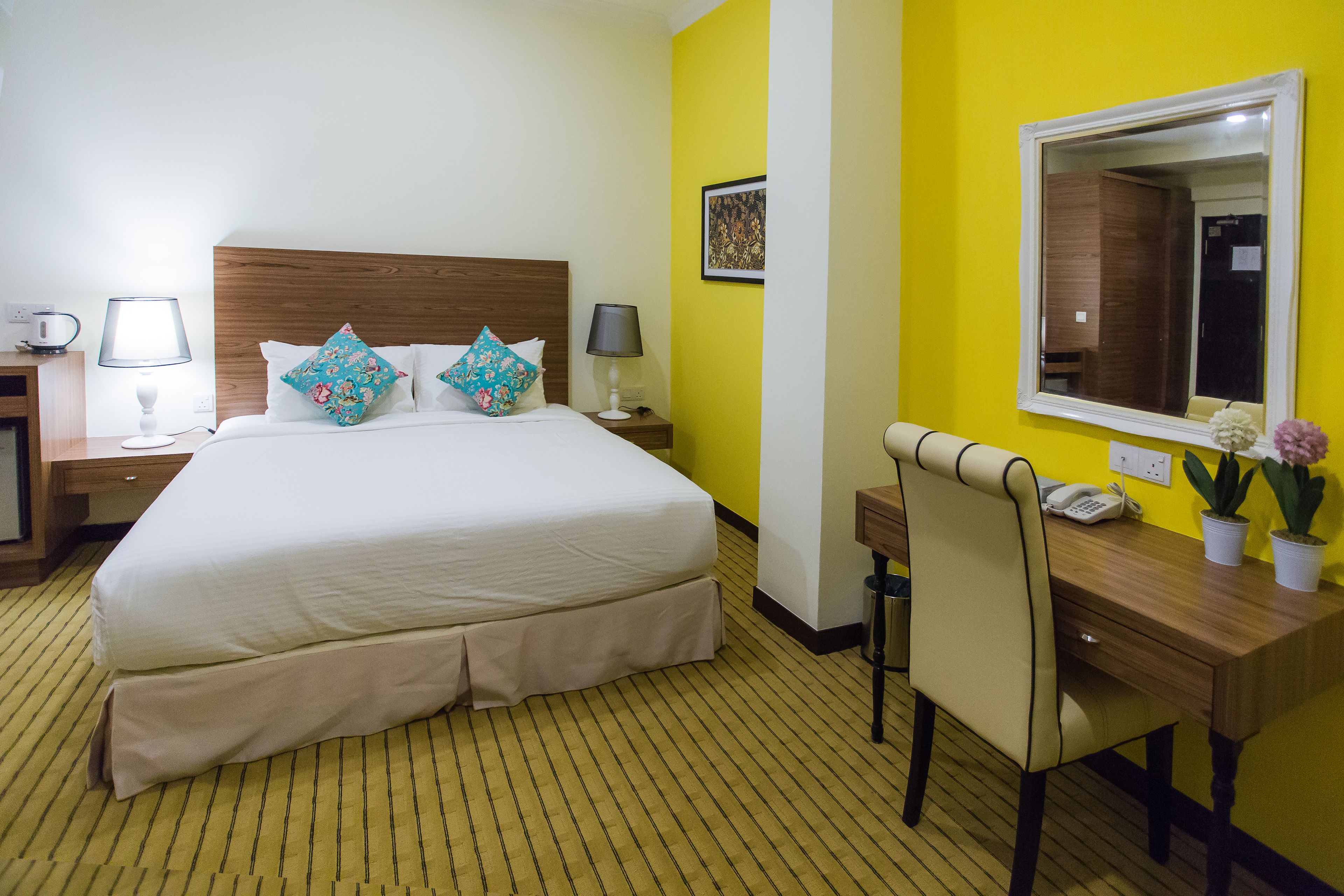 Quarto de hotel com uma cama grande, uma mesa com cadeira, um espelho e uma pequena mesa com abajur.