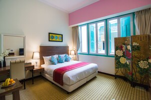 Quarto de hotel com cama, luminárias de cabeceira, escrivaninha com cadeira, um cesto de frutas e janela com cortinas.