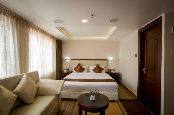 Quarto de hotel com cama, sofá, mesa de cabeceira com um vaso e porta de madeira.
