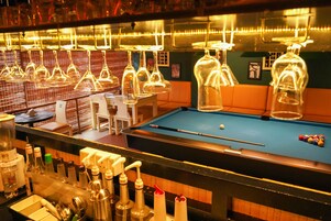 Uma área de bar com mesa de bilhar, banquetas e diversos tipos de garrafas e copos.