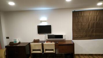 Quarto de hotel com escrivaninha, duas cadeiras, televisão e janela com cortinas.