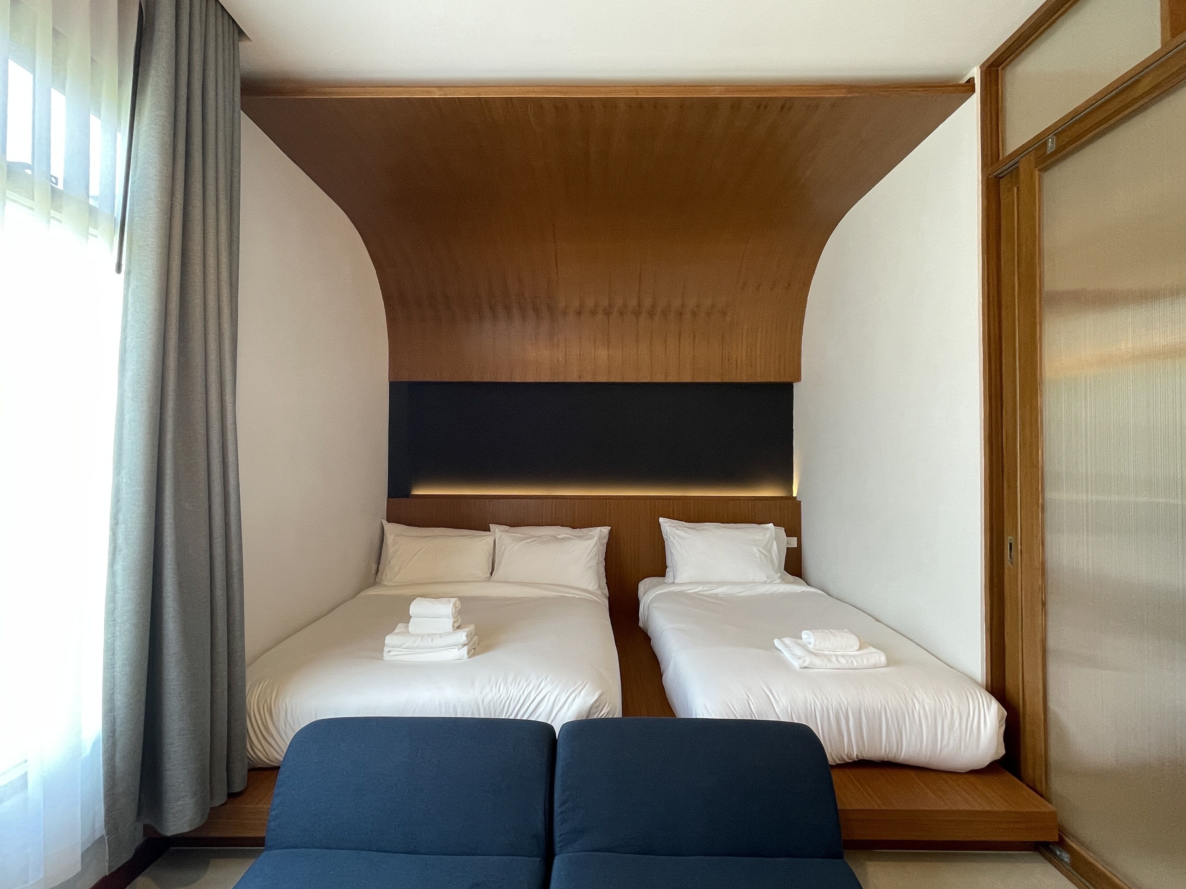 Quarto de hotel com duas camas, cabeceira de madeira e uma janela grande com cortinas.