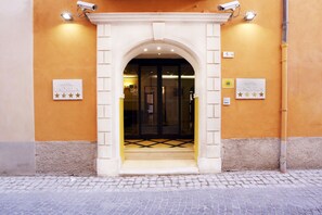 Entrada de um hotel com arco branco, porta de vidro e chão de azulejos. Há placas na parede com o nome do hotel e sua classificação por estrelas.