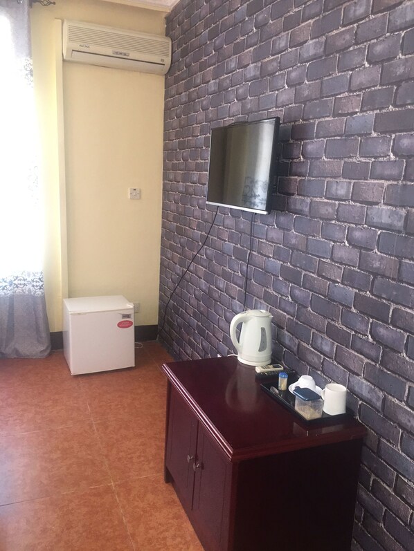 Quarto com parede de tijolos, televisão montada, pequeno frigorífico branco, armário de madeira com chaleira e chávenas, e chão em azulejos.
