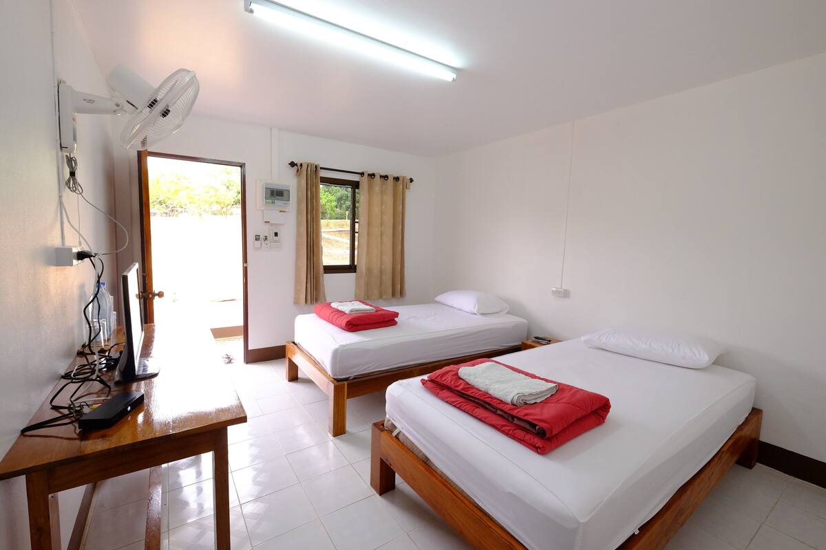 Quarto de hotel com duas camas de madeira, uma mesa com computador, ventoinha fixada na parede e janela com cortinas.