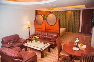Quarto de hotel com sofá de couro marrom, mesa de centro de madeira e mesa de jantar com cadeiras. Há uma luminária, um vaso com flores e uma garrafa sobre a mesa de jantar.