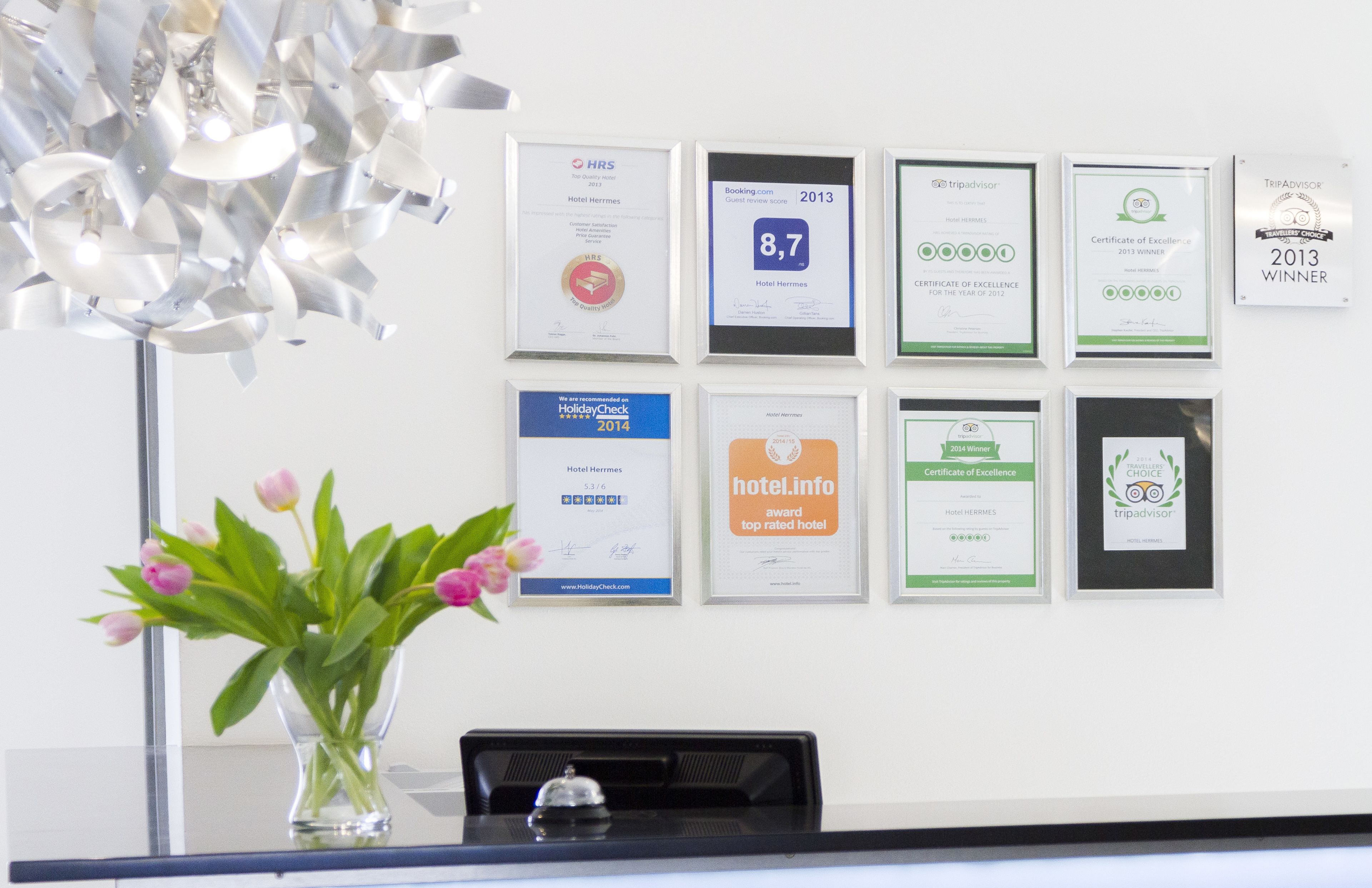 Uma parede exibindo diversos prêmios e certificados de um hotel, incluindo o prêmio de Vencedor TripAdvisor 2013 e o selo de Hotel com Melhor Avaliação HolidayCheck 2014.