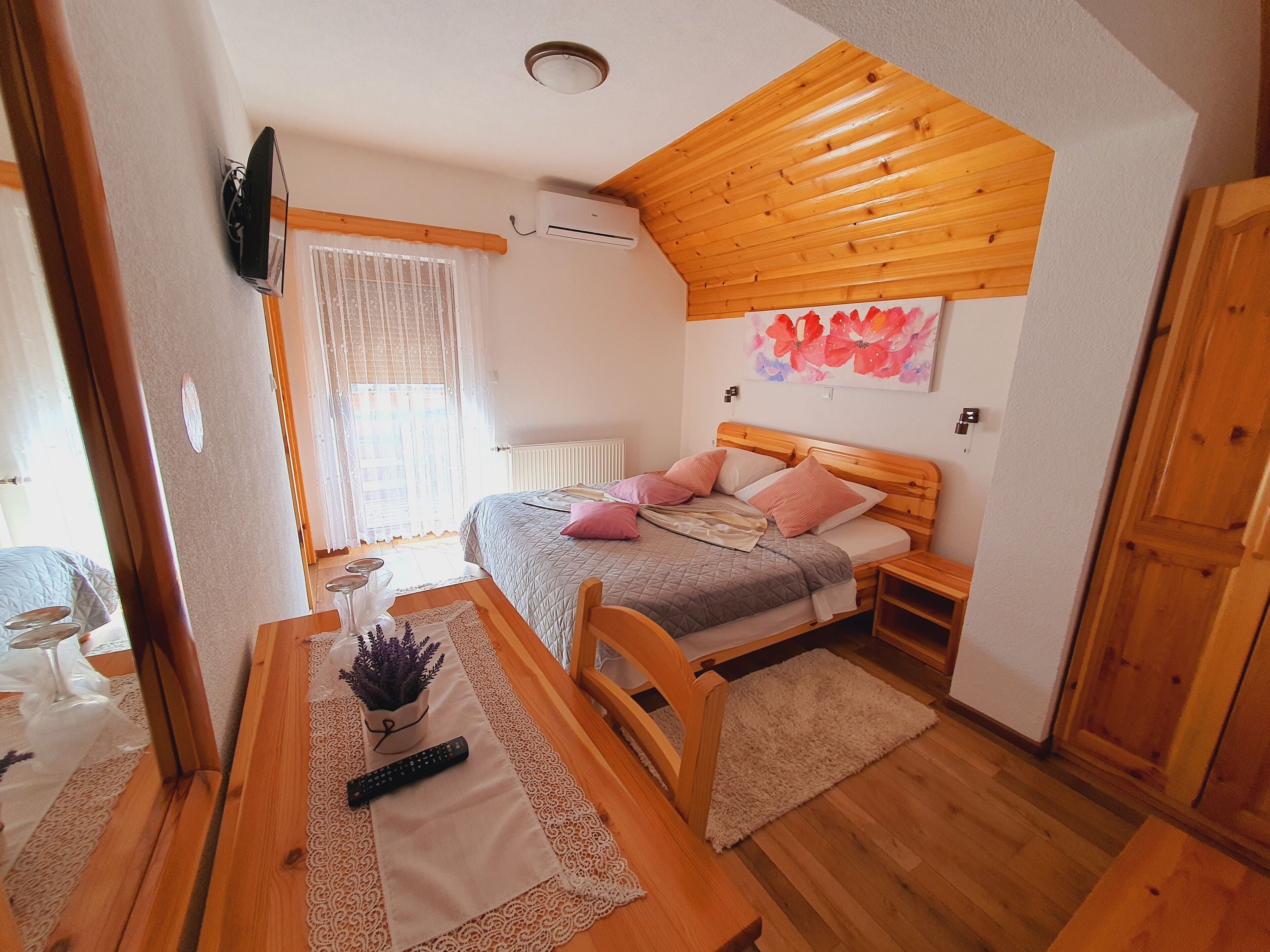 Quarto aconchegante com teto de madeira, cama com roupa de cama rosa e cinza, mesa de cabeceira pequena e televisão presa na parede.