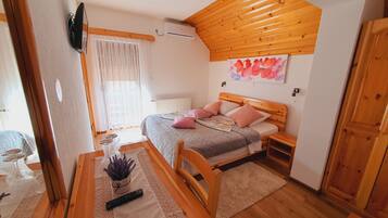 Quarto aconchegante com teto de madeira, cama com roupa de cama rosa e cinza, mesa de cabeceira pequena e televisão presa na parede.