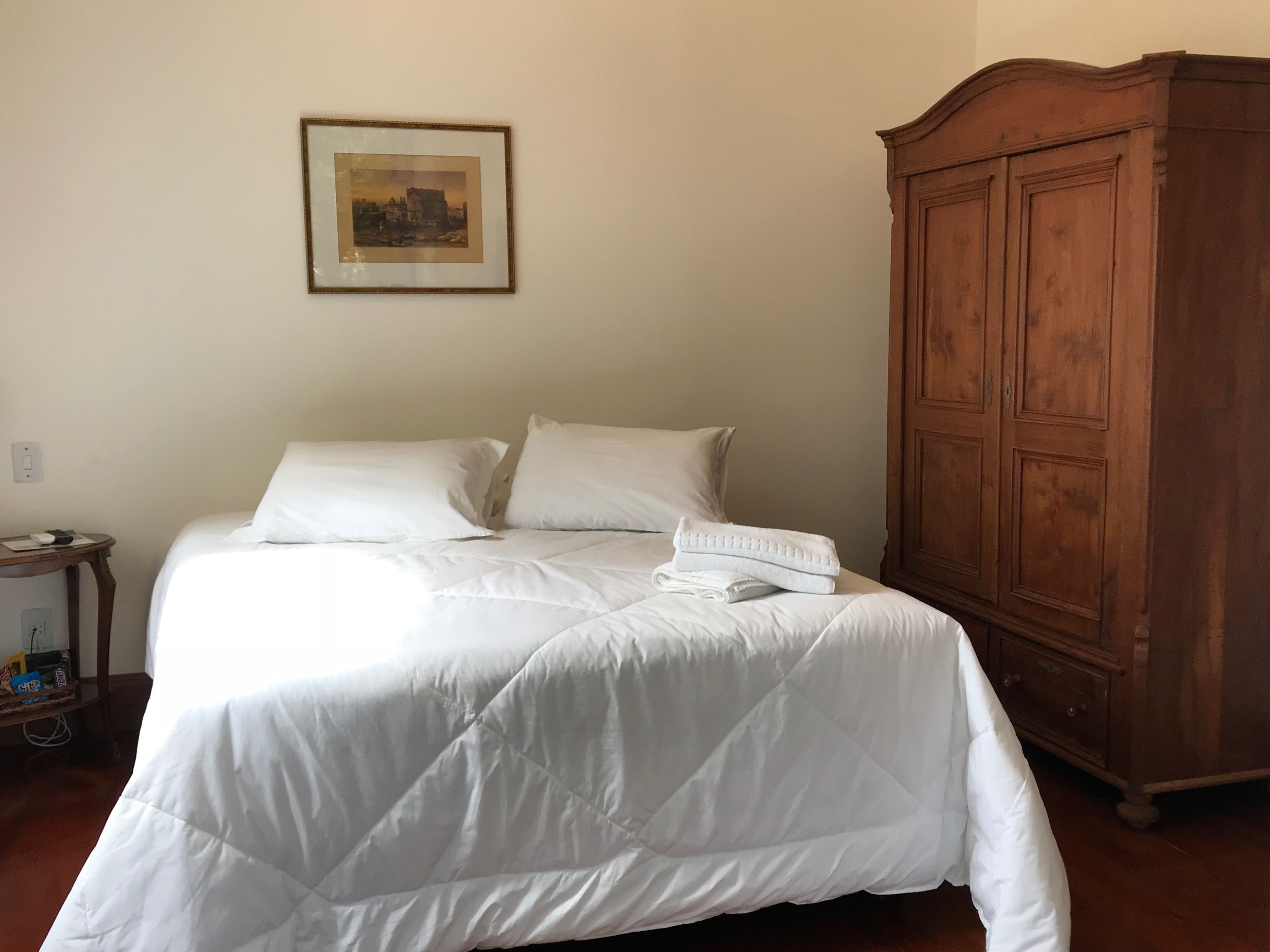 Uma cama bem arrumada com lençóis e almofadas brancos, um guarda-roupa de madeira e um quadro na parede.